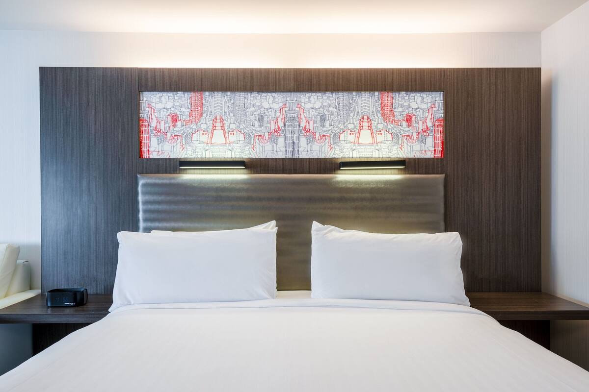 Quarto de hotel moderno com uma cama grande, duas almofadas brancas, uma mesa de cabeceira com relógio digital e uma obra de arte na parede.