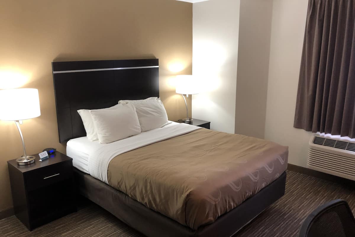 Quarto de hotel com uma cama, duas mesas de cabeceira com luminárias, ar condicionado na parede e uma janela com cortinas.