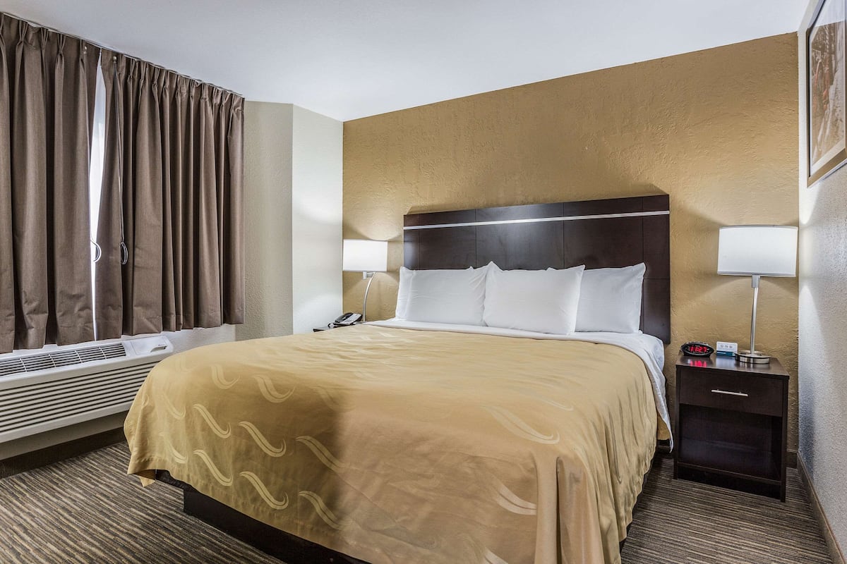 Quarto de hotel com uma cama grande, mesa de cabeceira com abajur, criado-mudo e uma janela com cortinas.