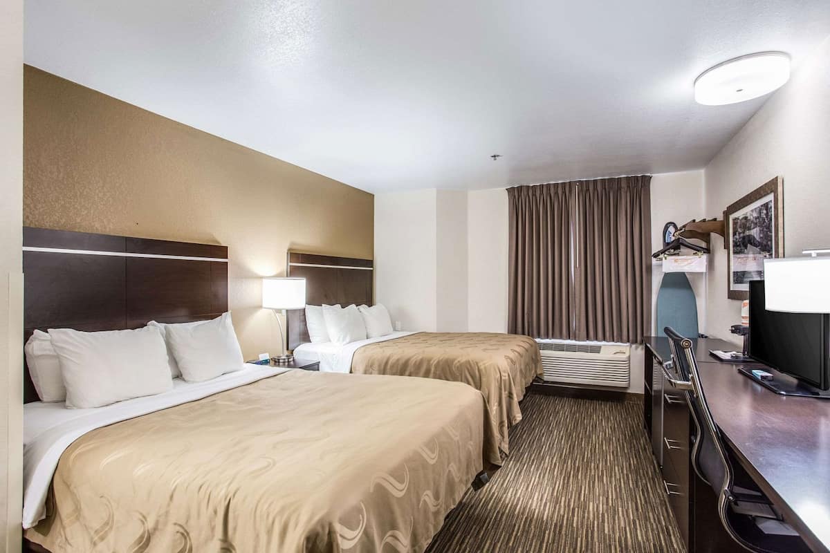 Quarto de hotel com duas camas, uma escrivaninha, uma cadeira, televisão, luminária e uma janela grande com cortinas.