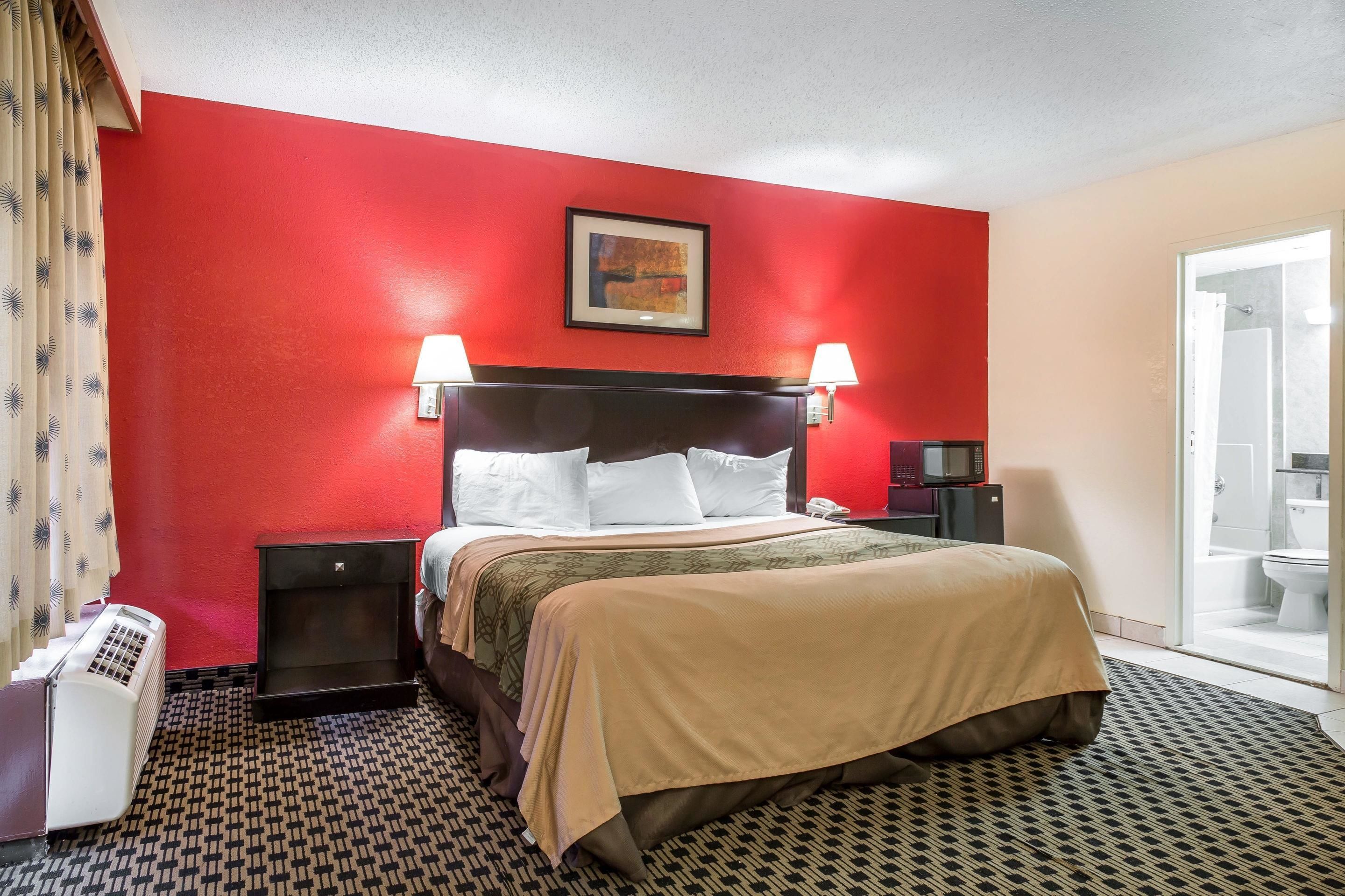 Quarto de hotel com parede de destaque vermelha, cama com roupa de cama branca, duas luminárias de cabeceira, um aquecedor e vista para o banheiro.