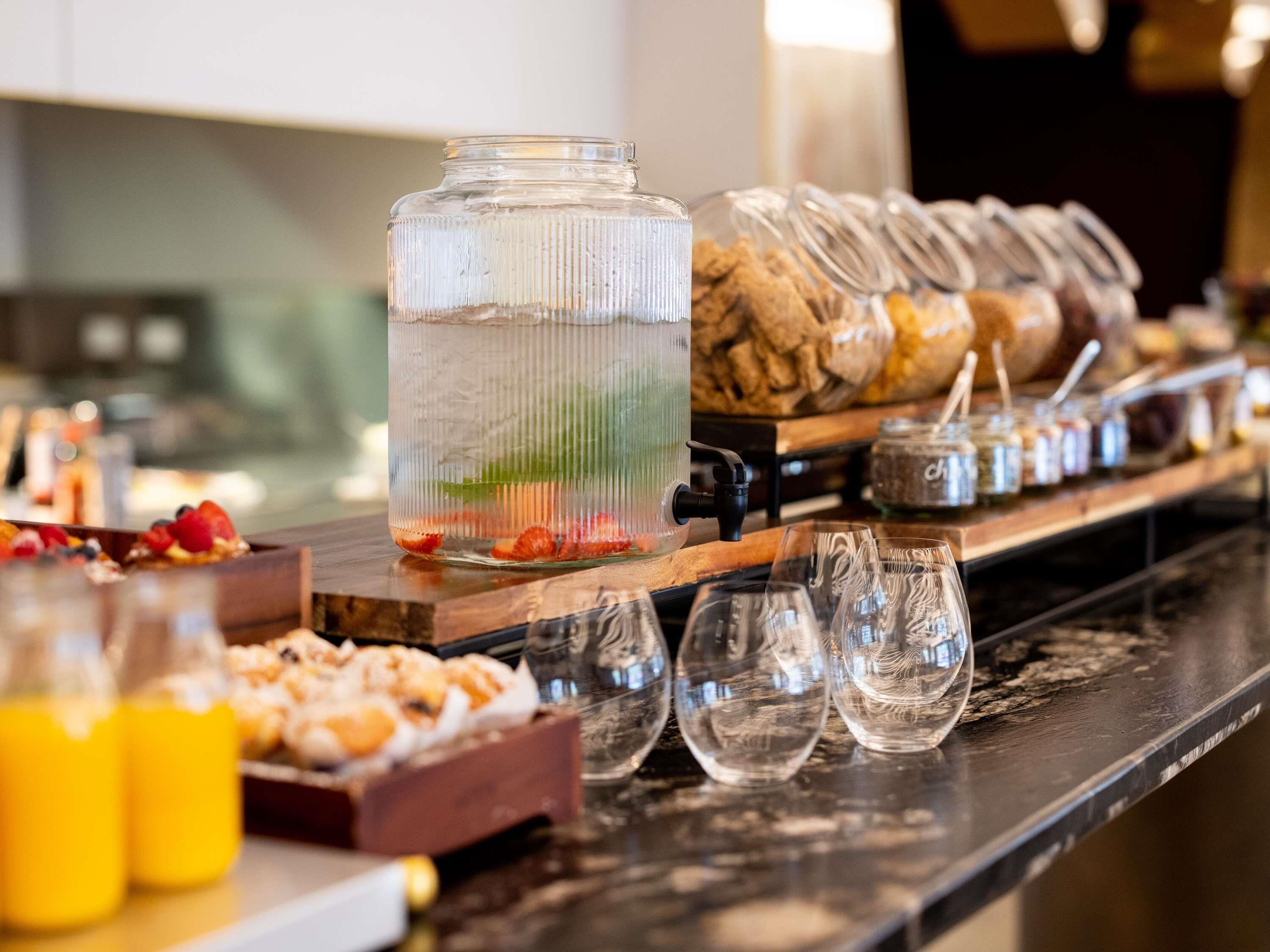 Disposição de buffet com um grande dispensador de água, diversos alimentos e copos vazios em um balcão.