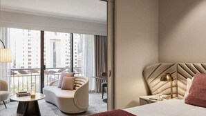 Quarto de hotel com uma cama, um sofá, uma mesa pequena, uma luminária e vista para prédios por grandes janelas.