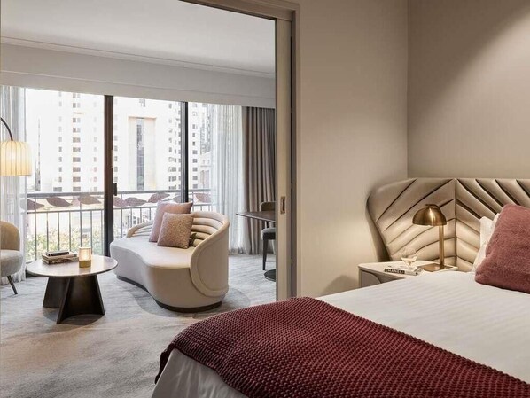 Quarto de hotel com uma cama, um sofá, uma mesa pequena, uma luminária e vista para prédios por grandes janelas.
