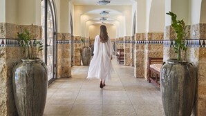 Um corredor com vasos grandes, uma mulher vestida de branco e um banco.
