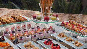 Uma mesa de buffet com diversos doces e bebidas, ao fundo arquitetura tradicional e palmeiras.