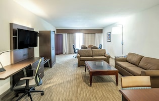 Quarto de hotel com televisão, escrivaninha, sofá, mesa de centro e uma grande janela com cortinas.