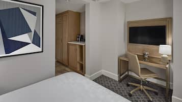 Quarto de hotel com cama, escrivaninha, cadeira, televisão e um quadro abstrato emoldurado.