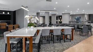 Uma zona de refeições de hotel moderna, com mesas de mármore, cadeiras estofadas cinzentas e uma planta em vaso central.