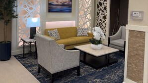 Um saguão de hotel moderno com um sofá, poltronas, uma mesinha de centro, painéis decorativos, um quadro e uma planta em vaso.