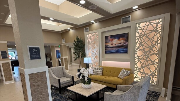 Um hall de hotel com um sofá, cadeiras, uma mesa de centro, uma planta decorativa e um quadro na parede.