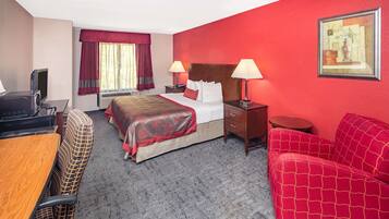 Quarto de hotel com cama, duas mesas de cabeceira com luminárias, escrivaninha com computador, sofá vermelho e piso coberto por carpete.