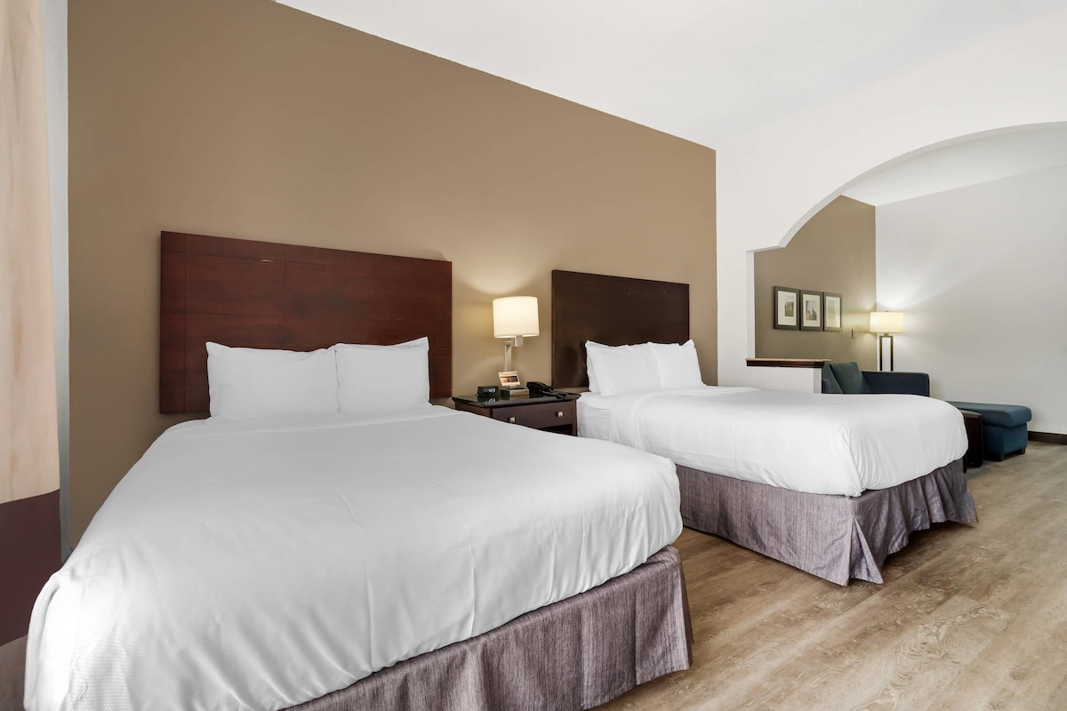 Quarto de hotel com duas camas, cabeceiras de madeira, mesa de cabeceira com abajur e um pequeno espaço de estar com um banco.