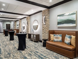 Um lobby de hotel com um sofá de couro marrom, mesas redondas, um balcão de recepção e uma parede de destaque em pedra.