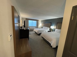 Quarto de hotel com duas camas, uma mesa de trabalho com computador, uma janela com cortinas e ar condicionado fixado na parede.
