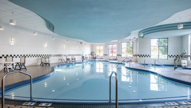 Piscina interior com chão em azulejo, cadeiras de descanso e ampla vista para o exterior através de grandes janelas.