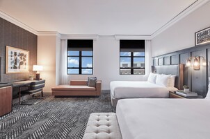 Quarto de hotel com duas camas, uma escrivaninha, um sofá e uma janela grande.