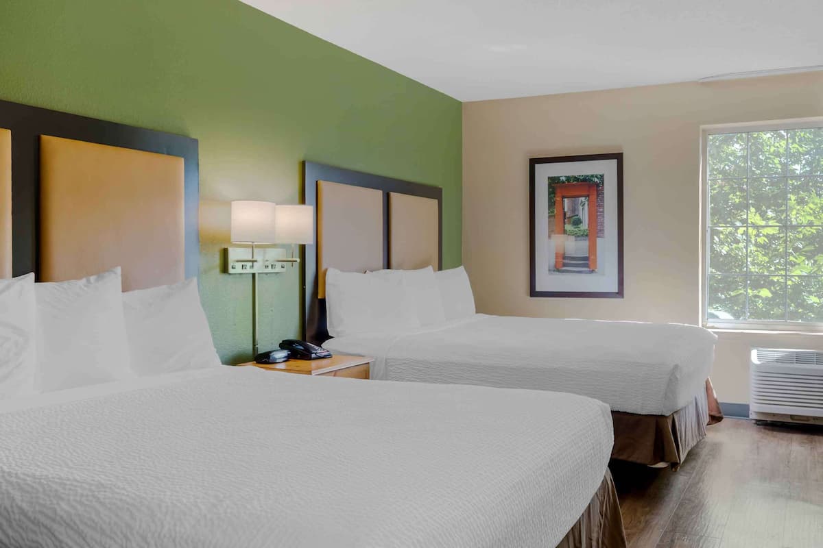 Quarto de hotel com duas camas, uma mesa de cabeceira com abajur, um quadro na parede e uma janela com vista para árvores.