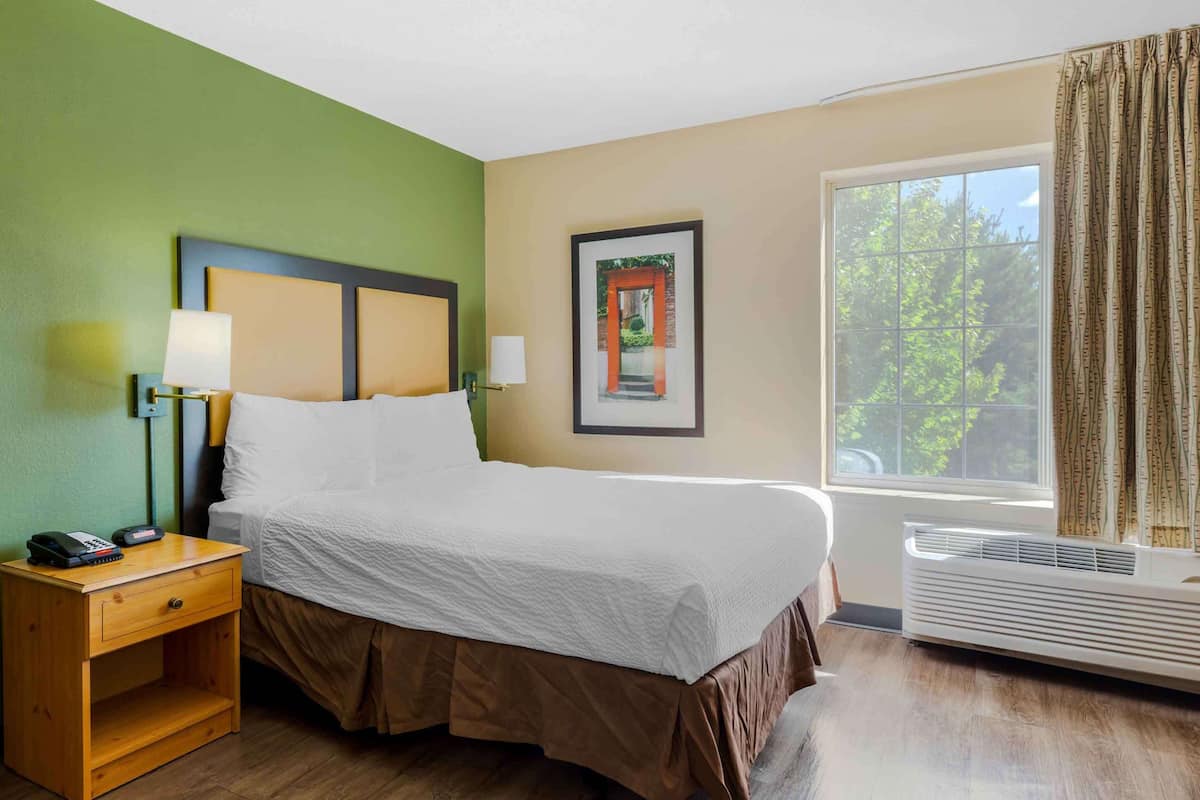 Quarto de hotel com cama, mesa de cabeceira, luminária, ar condicionado e janela com cortinas.