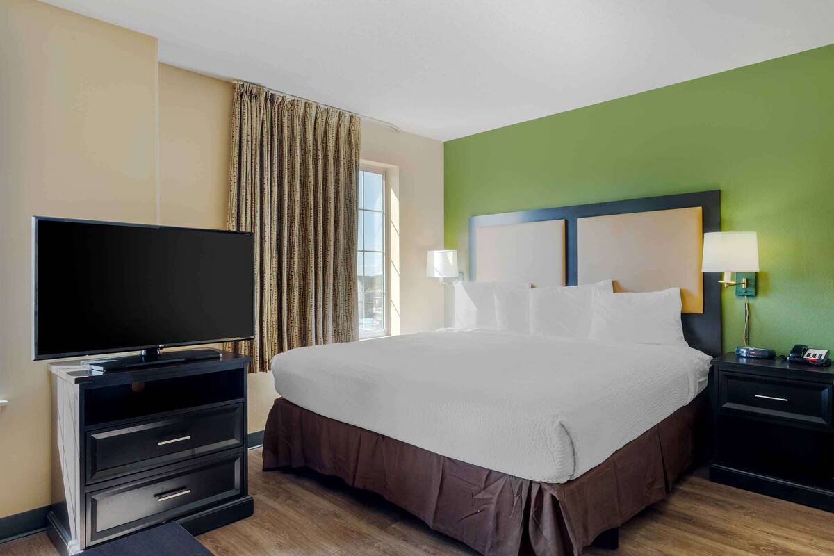Quarto de hotel com uma cama grande, televisão de tela plana, mesa de cabeceira com abajur e janela com cortinas.