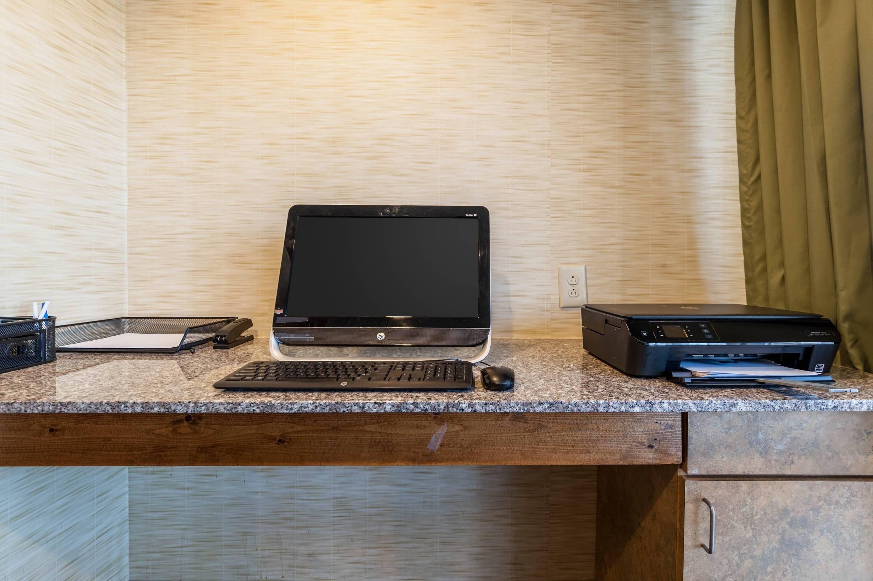 Um espaço de trabalho com um laptop, teclado, rato e impressora sobre uma escrivaninha de madeira, em frente a uma parede com textura.
