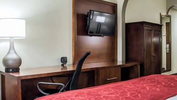 Quarto de hotel com escrivaninha de madeira, televisão na parede, luminária, cadeira, armário e colcha vermelha.