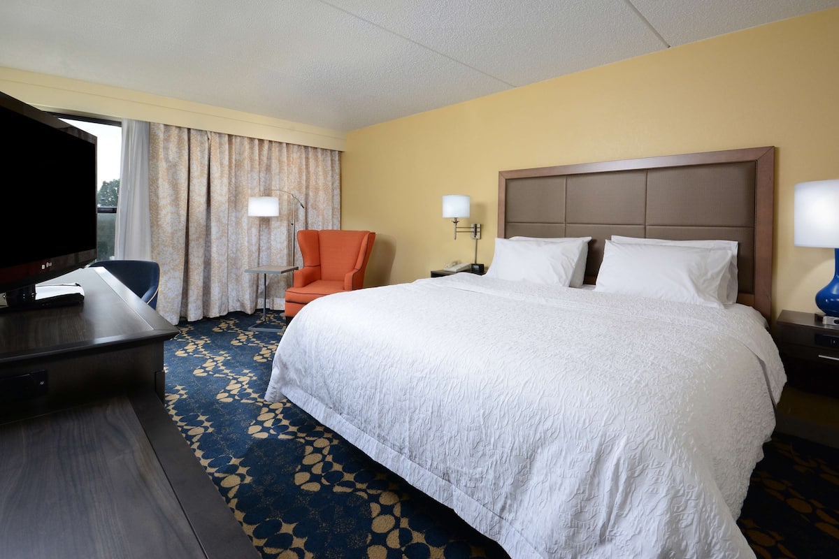 Quarto de hotel com uma cama grande, uma escrivaninha com televisão, uma cadeira laranja e um abajur.