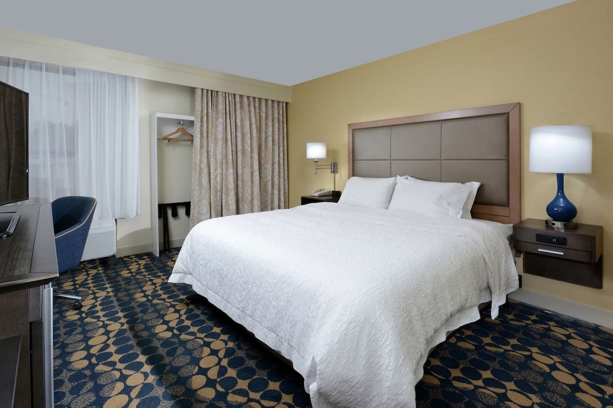 Quarto de hotel com uma cama grande, uma escrivaninha, uma cadeira, uma mesa de cabeceira, um abajur e um guarda-roupa.