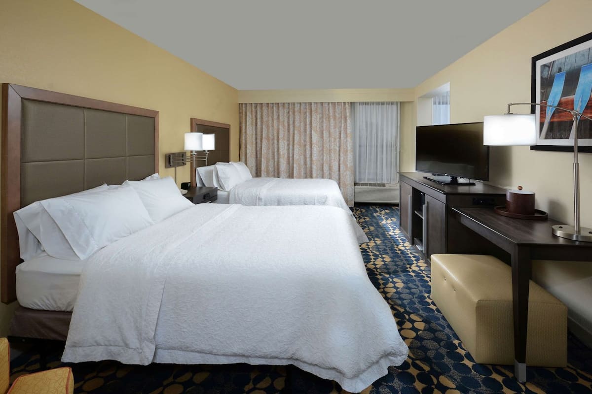Quarto de hotel com duas camas, uma escrivaninha, televisão e uma janela grande com cortinas.