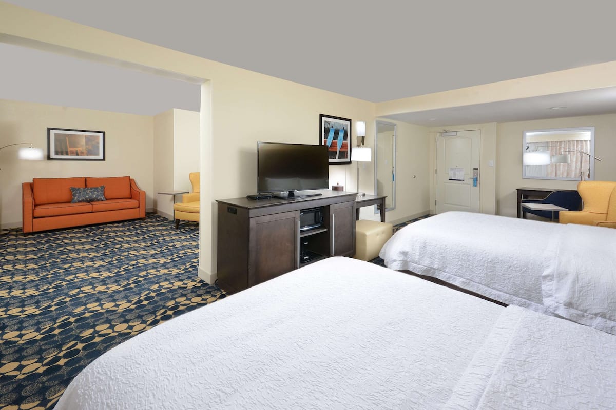 Quarto de hotel com duas camas, um sofá, uma escrivaninha com televisão e uma mesinha.
