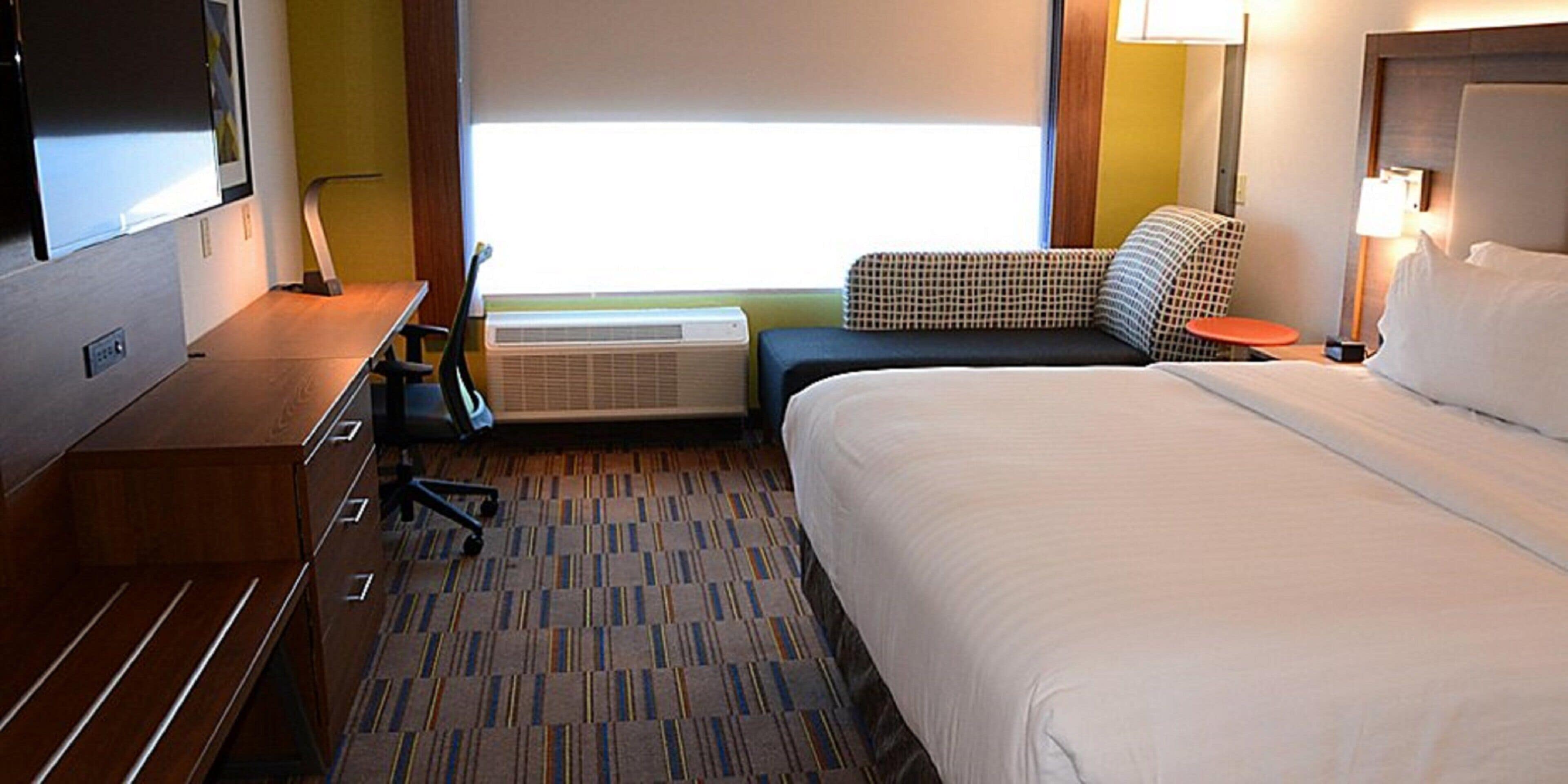 Quarto de hotel com cama, escrivaninha, ar condicionado, janela com persianas e luminária.