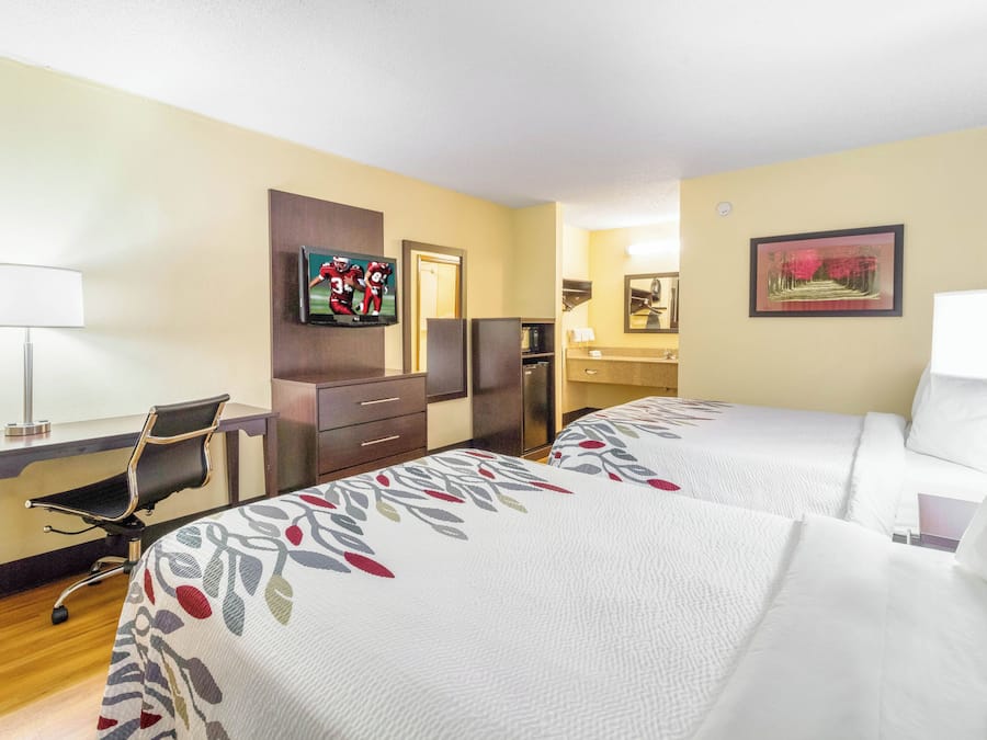 Quarto de hotel com duas camas, uma escrivaninha, uma cadeira, televisão, máquina de café e banheiro com espelho e porta-toalhas.