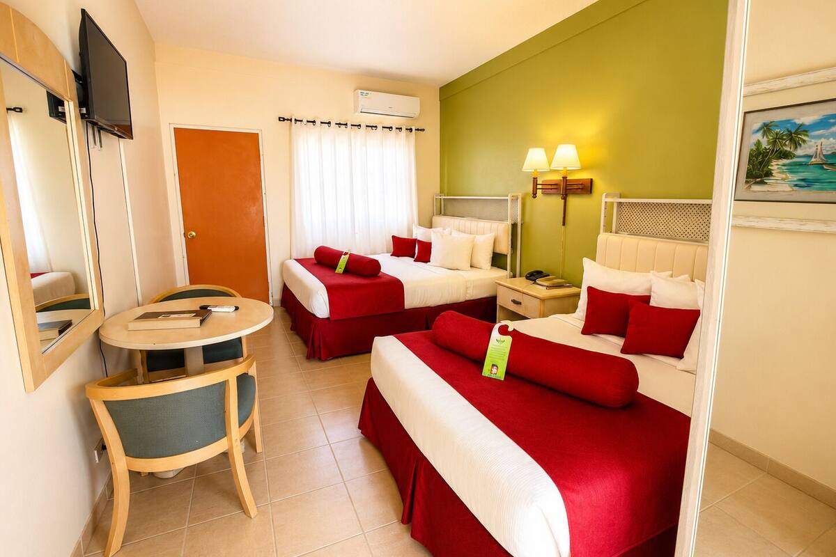 Quarto de hotel com duas camas, televisão, ar condicionado, escrivaninha com cadeira e um quadro na parede.
