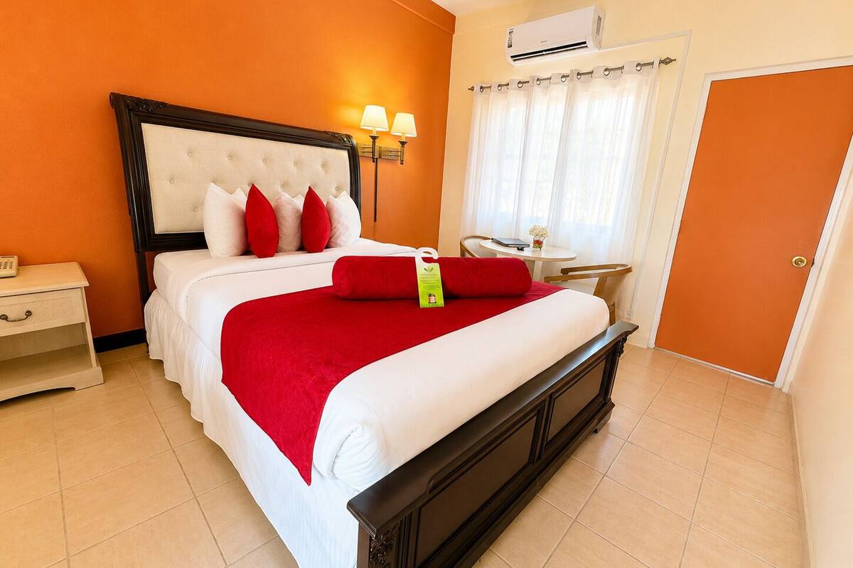 Quarto de hotel com cama, mesa de cabeceira, luminária, aparelho de ar condicionado e janela com cortinas.