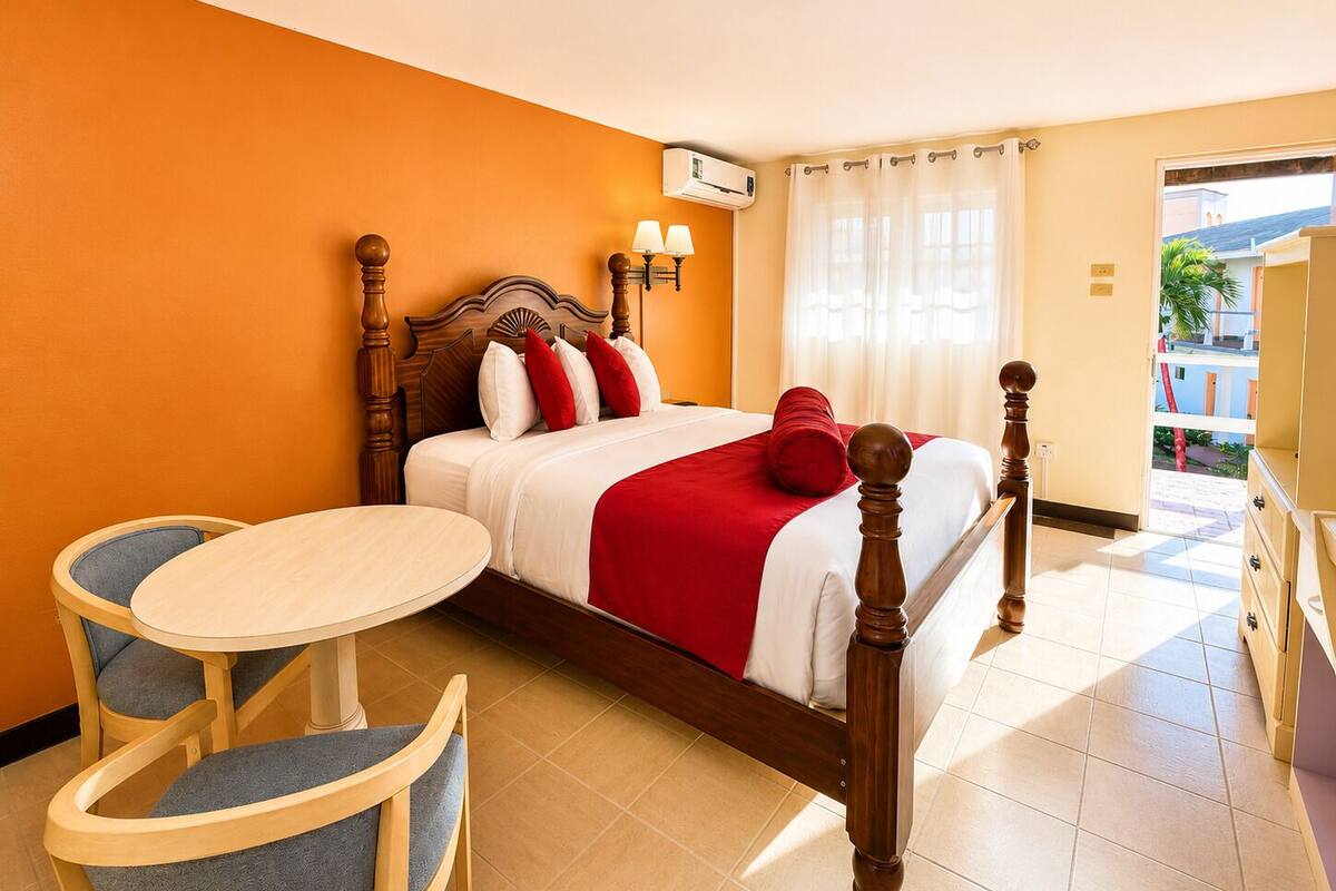Quarto de hotel com cama, mesa redonda pequena, duas cadeiras, janela com cortinas e unidade de ar condicionado.