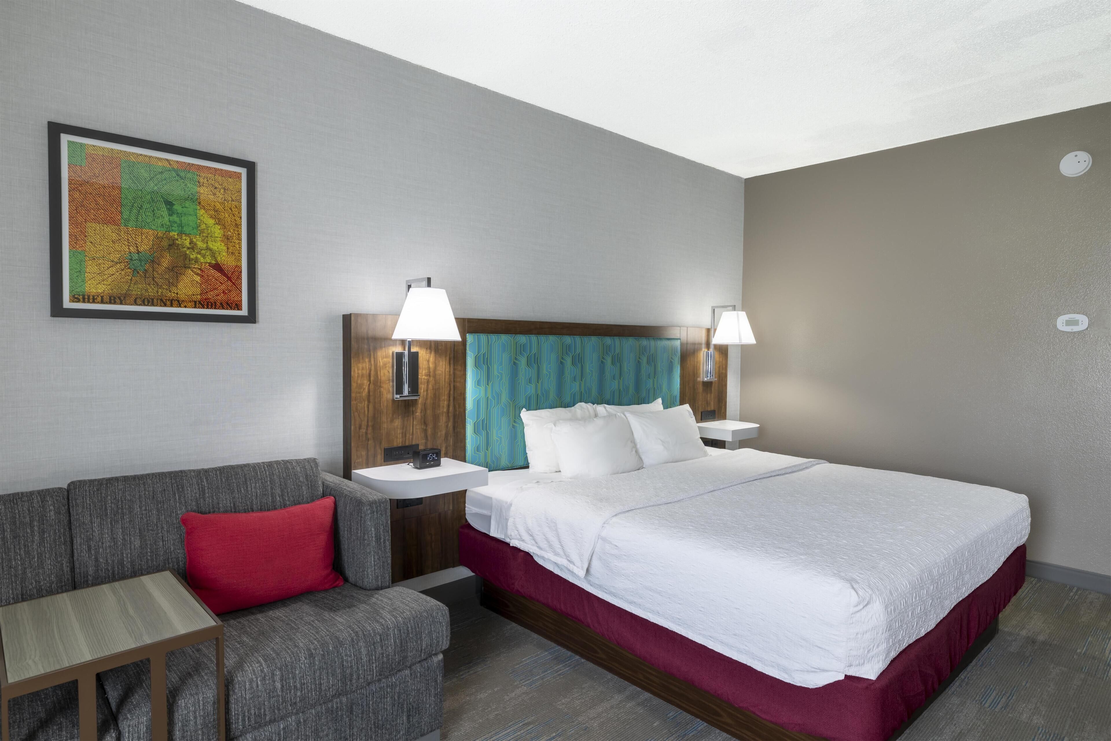 Quarto de hotel com uma cama grande, um sofá, duas mesinhas de cabeceira e uma obra de arte na parede.