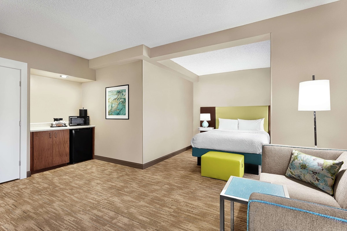 Quarto de hotel com cama, escrivaninha, cadeira, luminária e um quadro na parede.