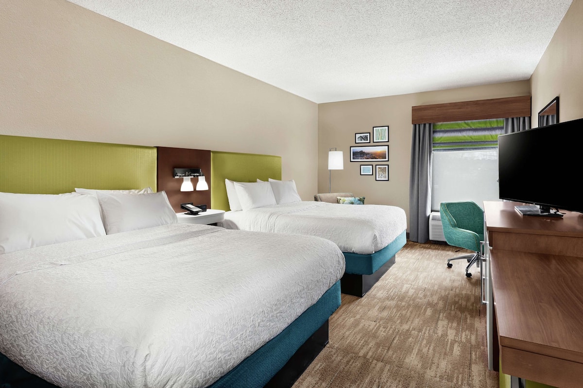 Quarto de hotel com duas camas, uma escrivaninha, uma cadeira, televisão e janela com cortinas.