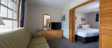 Quarto de hotel com sofá, cama, televisão, cômoda e porta de madeira que dá para outro espaço.