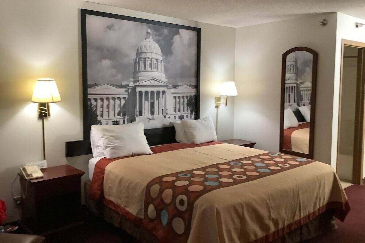 Quarto de hotel com uma cama grande, duas luminárias de cabeceira, uma escrivaninha com telefone e uma grande moldura com uma imagem de um edifício na parede.