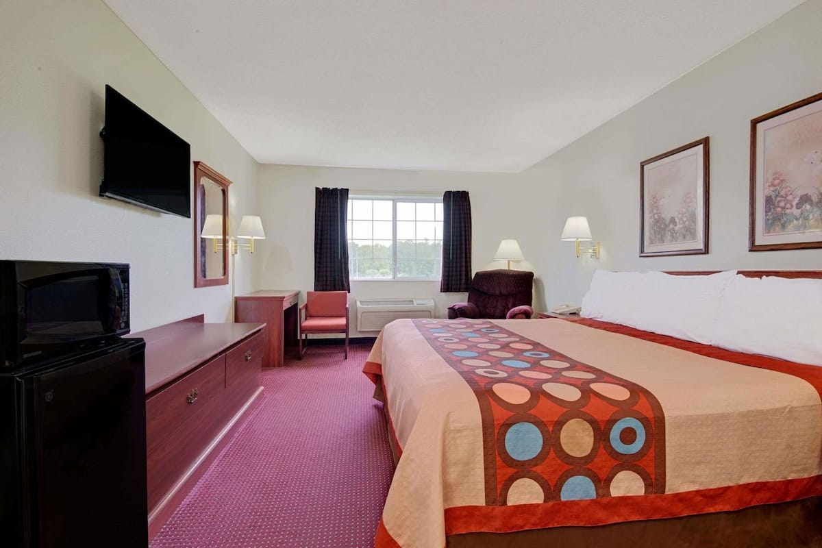 Quarto de hotel com cama, escrivaninha, cadeira, televisão e janela com cortinas.