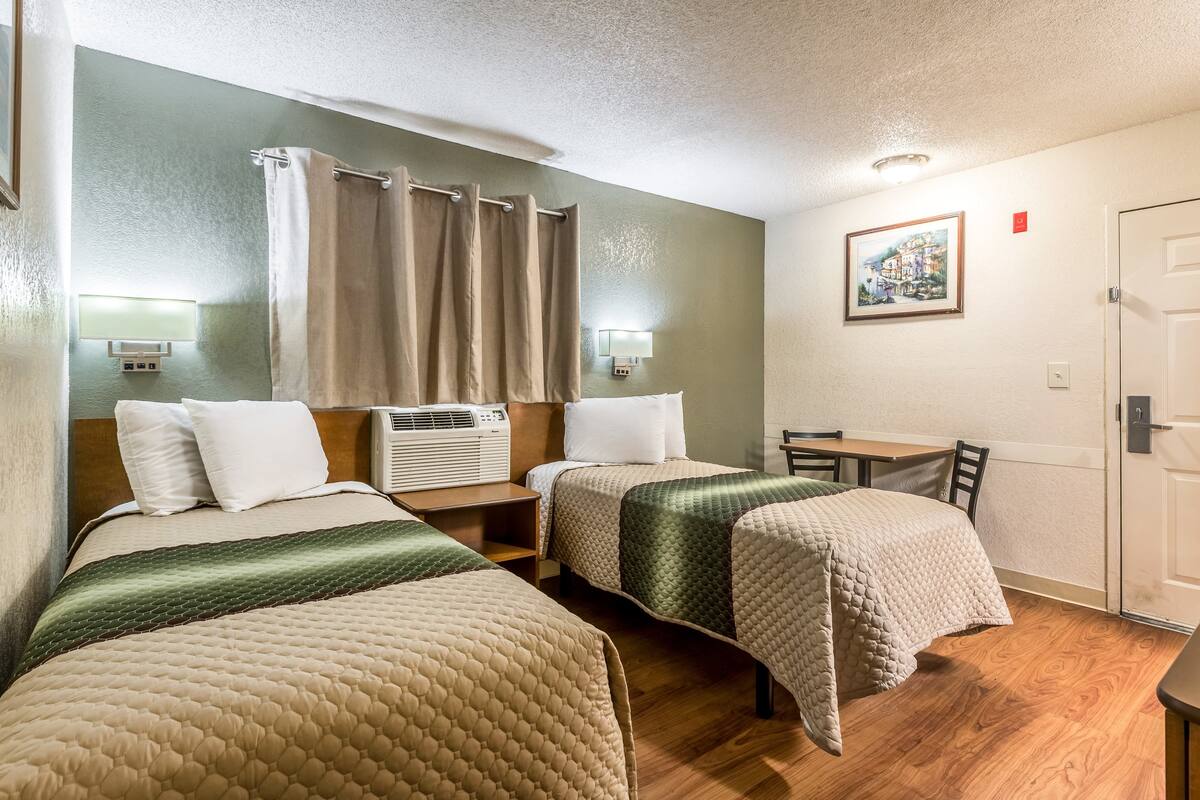 Quarto de hotel com duas camas, uma escrivaninha, uma cadeira, uma mesa, cortina de chuveiro, ar condicionado fixado na parede, luminária na parede e um quadro na parede.