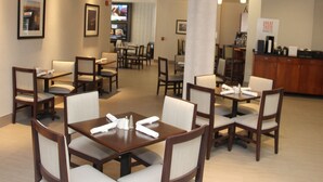 Uma área de refeições de hotel com várias mesas e cadeiras, uma receção e quadros nas paredes.