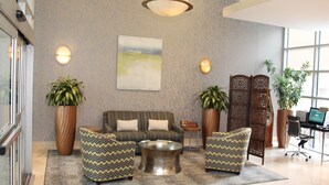 Um lobby moderno com um conjunto de sofás, mesa de centro, plantas em vasos e uma grande janela que oferece luz natural.