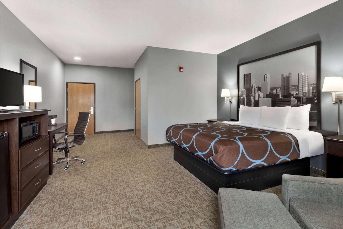Quarto de hotel com cama, escrivaninha com televisão, cadeira e arte de parede representando um horizonte urbano.
