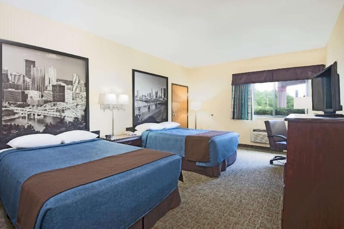 Quarto de hotel com duas camas, uma escrivaninha, uma cadeira, uma televisão e duas imagens emolduradas de paisagem urbana.