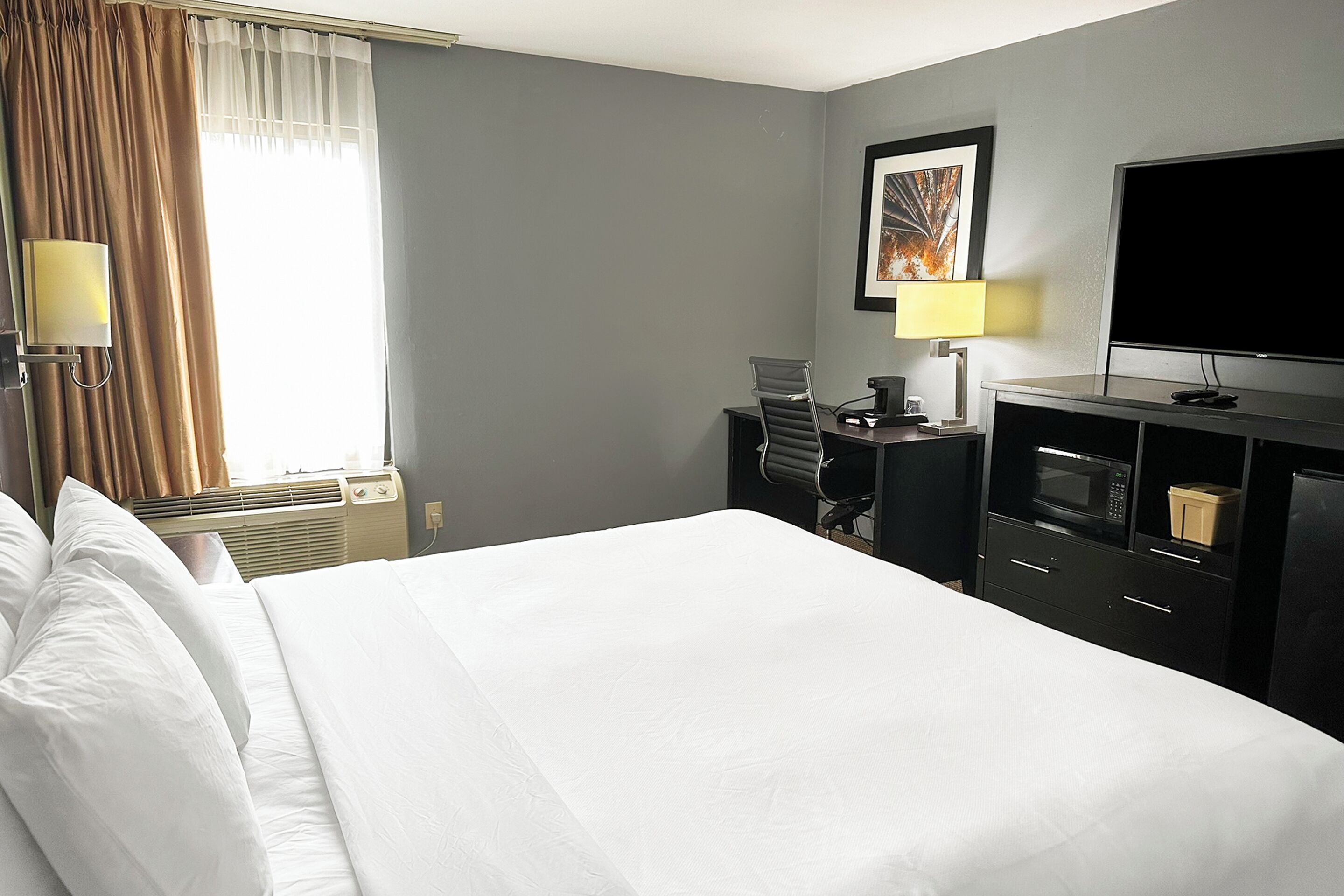 Quarto de hotel com uma cama grande, uma escrivaninha com cadeira, televisão de tela plana, micro-ondas e cafeteira.