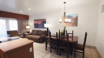 Quarto de hotel com cozinha compacta, área de refeições com mesa e cadeiras, sofá, luminária e um quadro na parede.