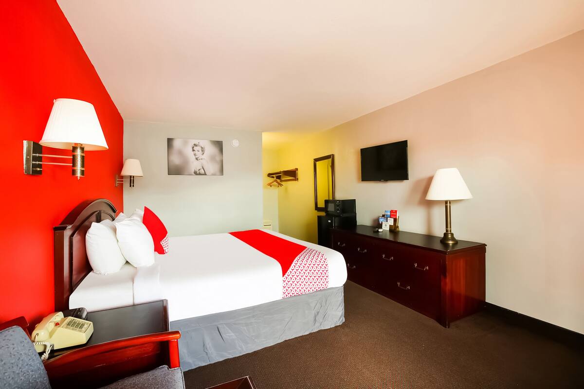 Quarto de hotel com cama, mesa de cabeceira, luminária, cômoda e televisão.