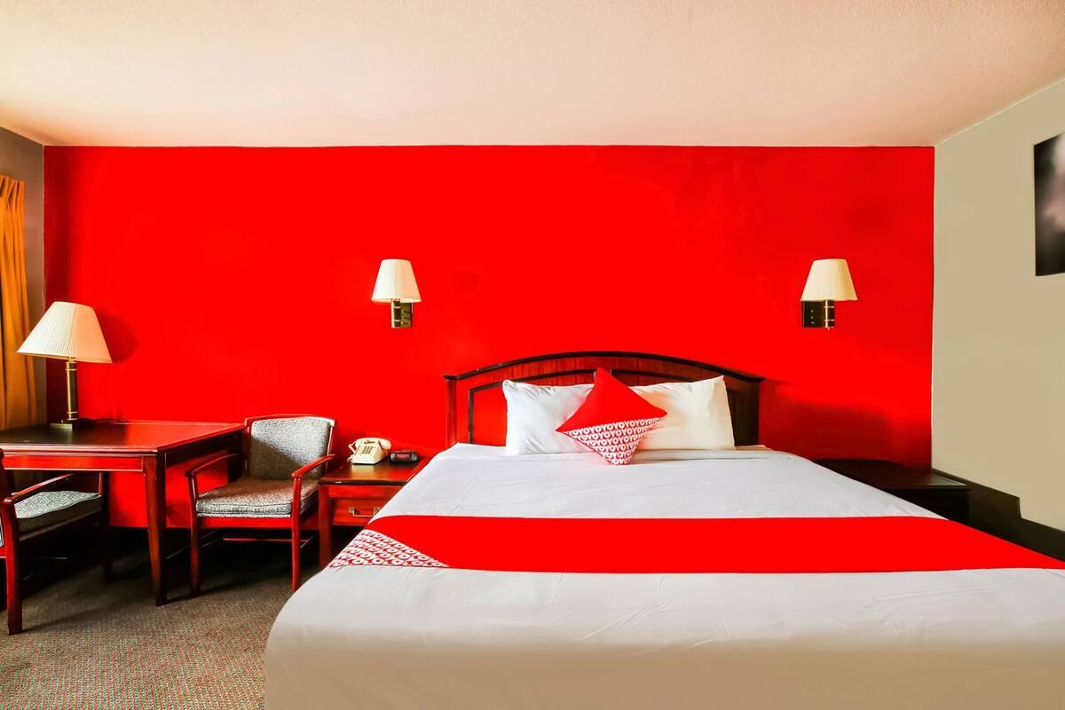 Quarto de hotel com parede vermelha, cama com roupa de cama branca, escrivaninha com abajur e uma cadeira.